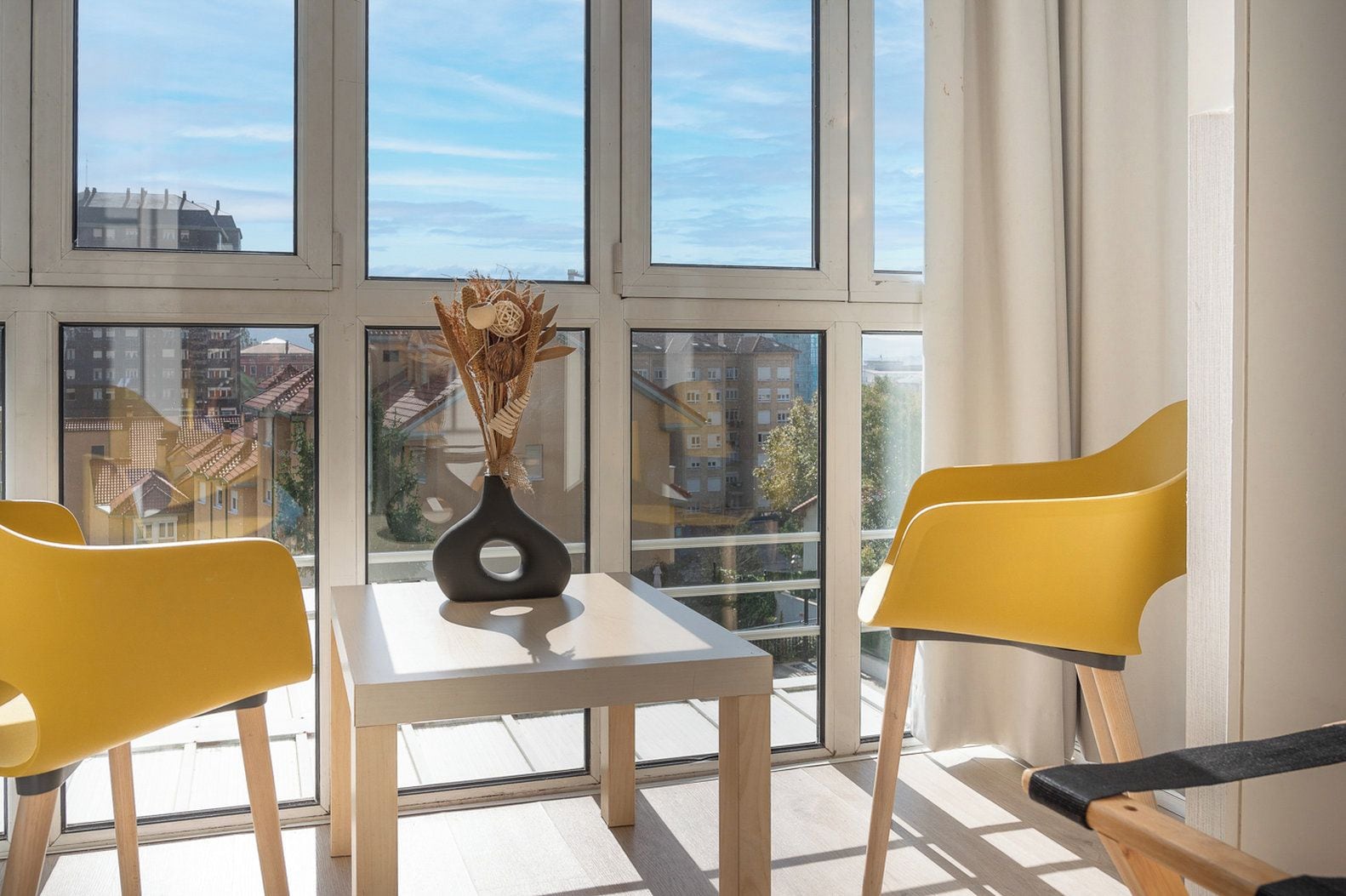 Quarto com mesa, duas cadeiras, um vaso com plantas secas e grandes janelas com vista para a cidade.