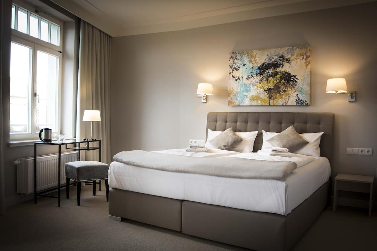 Quarto de hotel com uma cama grande, duas luminárias de cabeceira, um quadro na parede, uma escrivaninha com cadeira e um radiador.