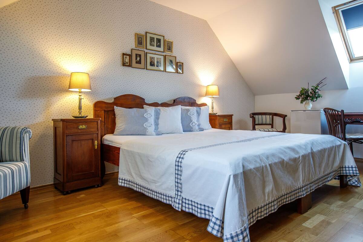 Quarto com cama, duas mesas de cabeceira com luminárias, cabeceira de madeira, uma cadeira e escrivaninha com planta.