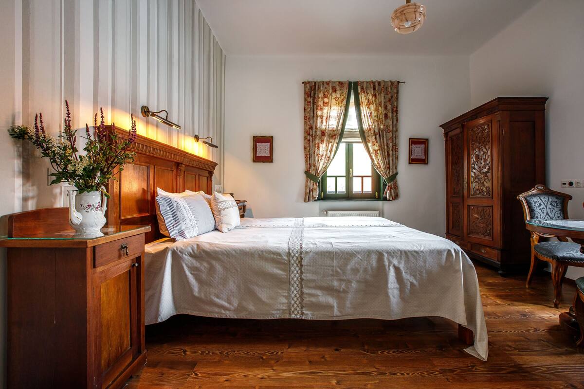 Quarto com cama grande, cabeceira de madeira, mesa de cabeceira, cômoda, cadeira e janela com cortinas.