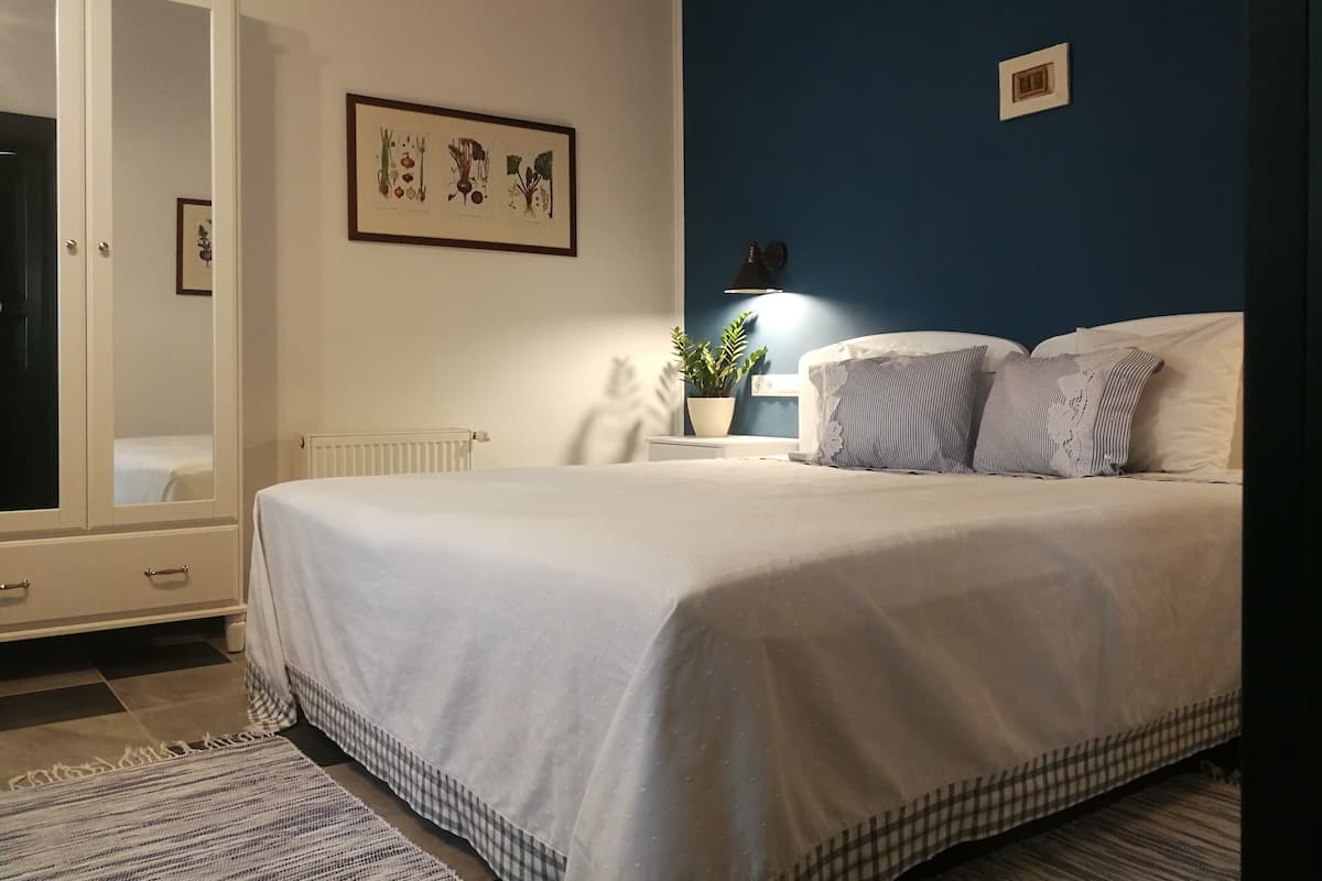 Quarto com cama, mesa de cabeceira, planta em vaso, ilustração botânica emoldurada e guarda-roupa branco.
