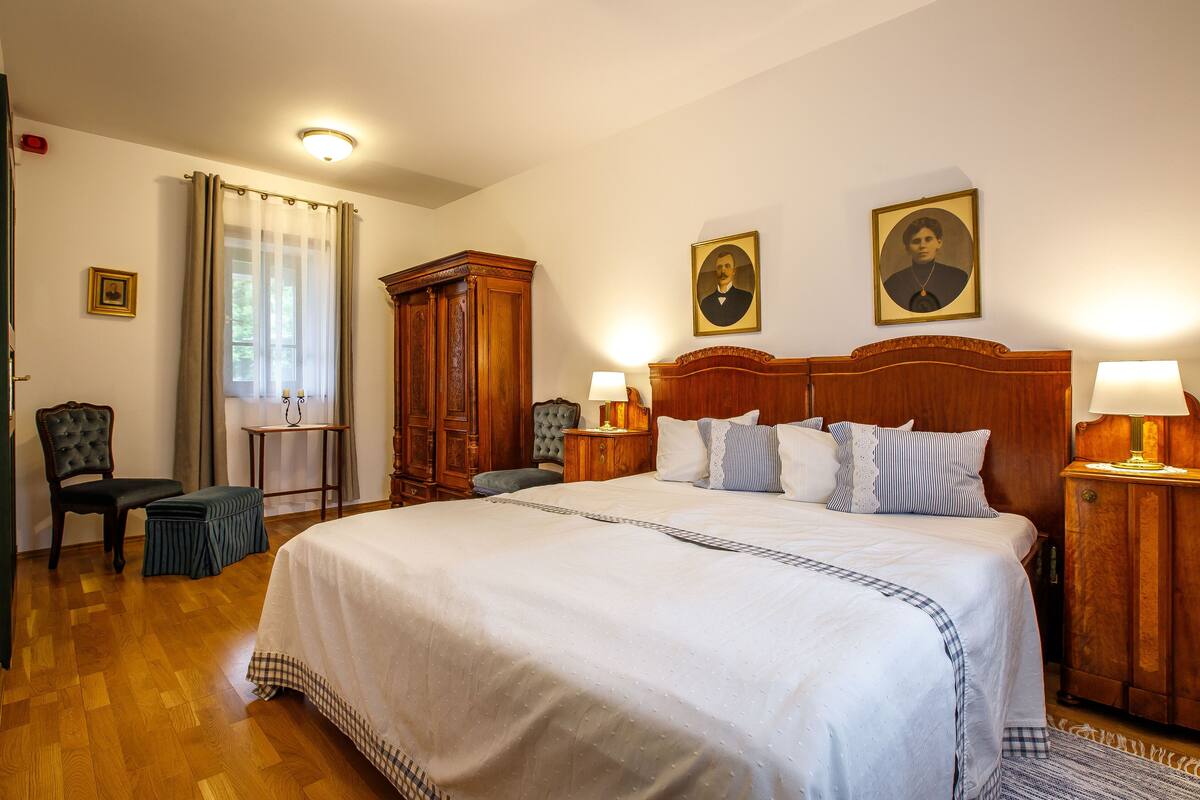 Quarto de hotel com uma cama grande, duas mesas de cabeceira com luminárias, um guarda-roupa de madeira, uma cadeira e uma pequena mesa.
