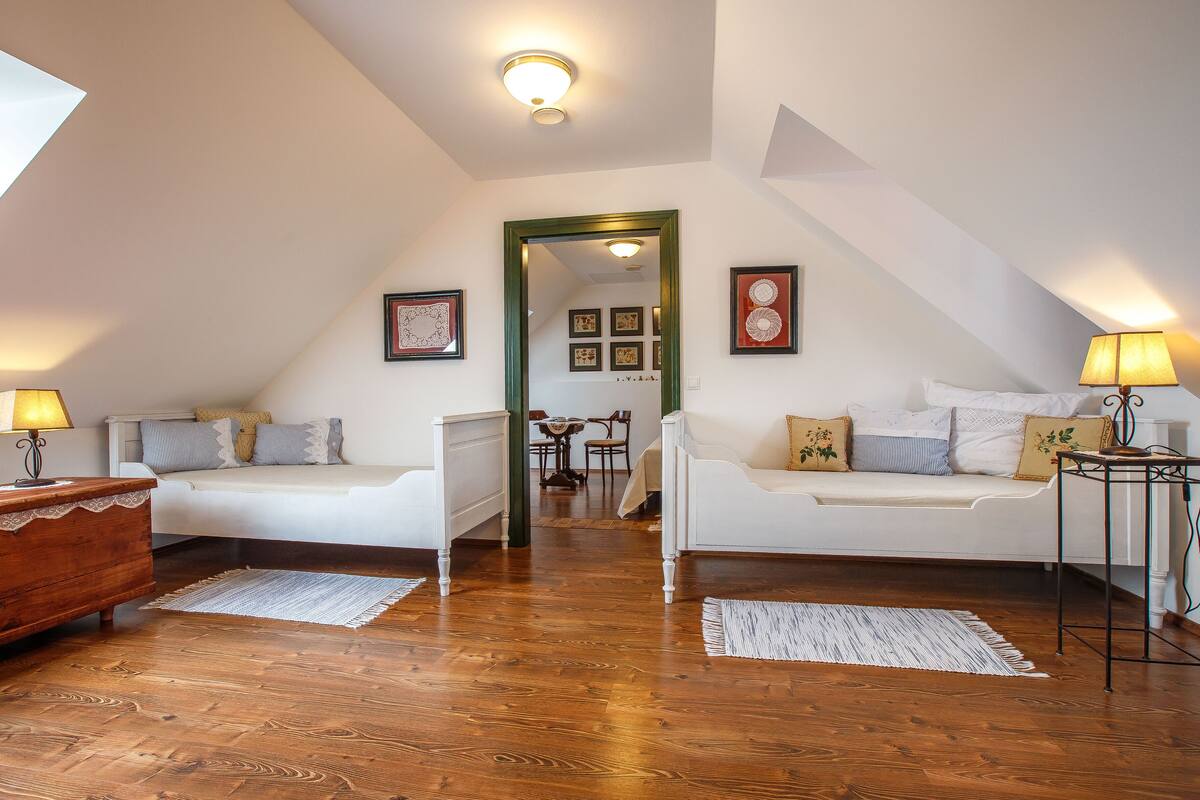 Um quarto de sótão aconchegante com piso de madeira, uma cama branca, um sofá branco e uma pequena mesa com abajur.