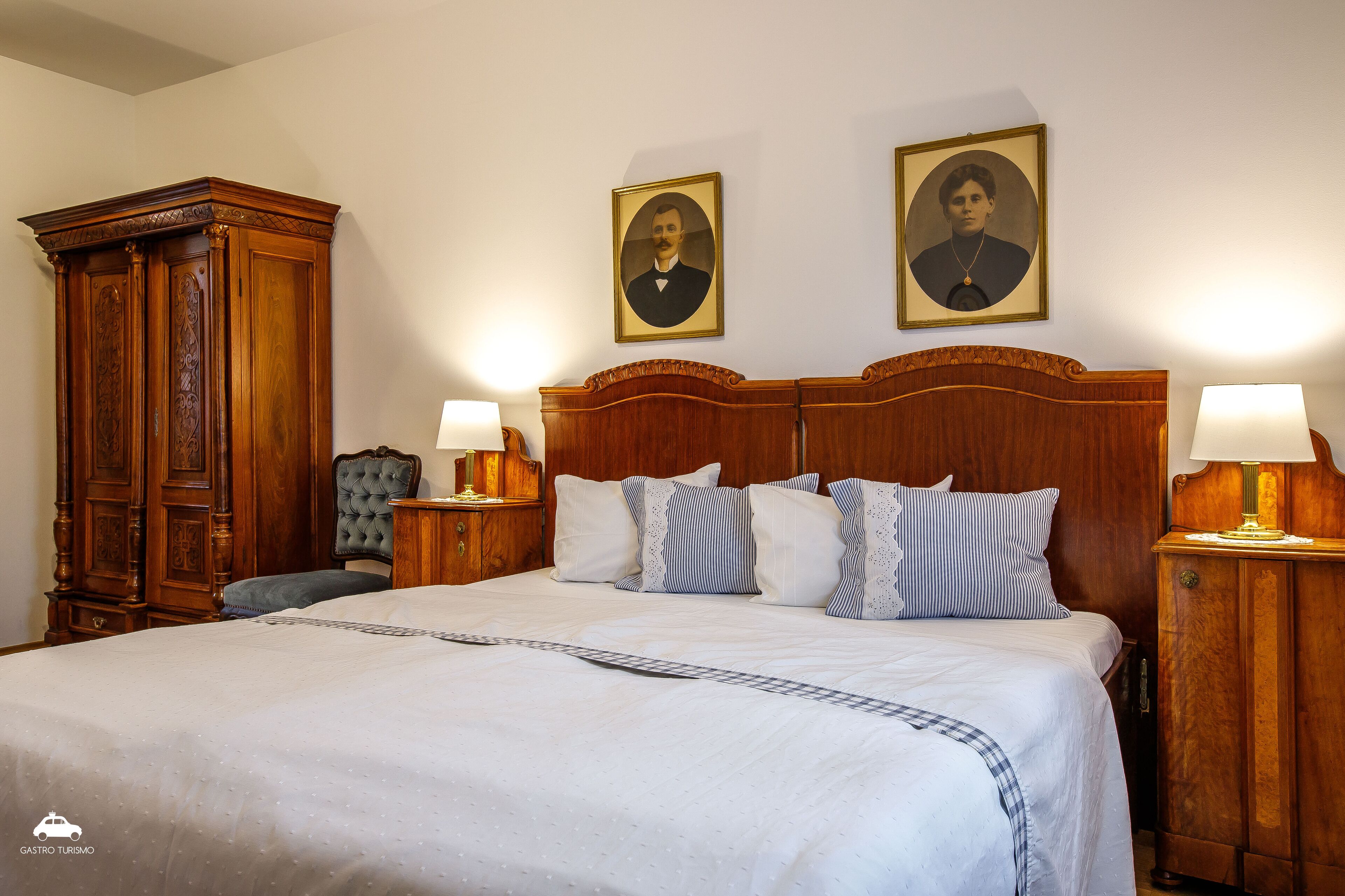 Uma cama bem arrumada com colcha branca e almofadas azuis e brancas listradas, com mesas de cabeceira de madeira com abajures de cada lado. Dois retratos emoldurados pendem na parede acima da cama.