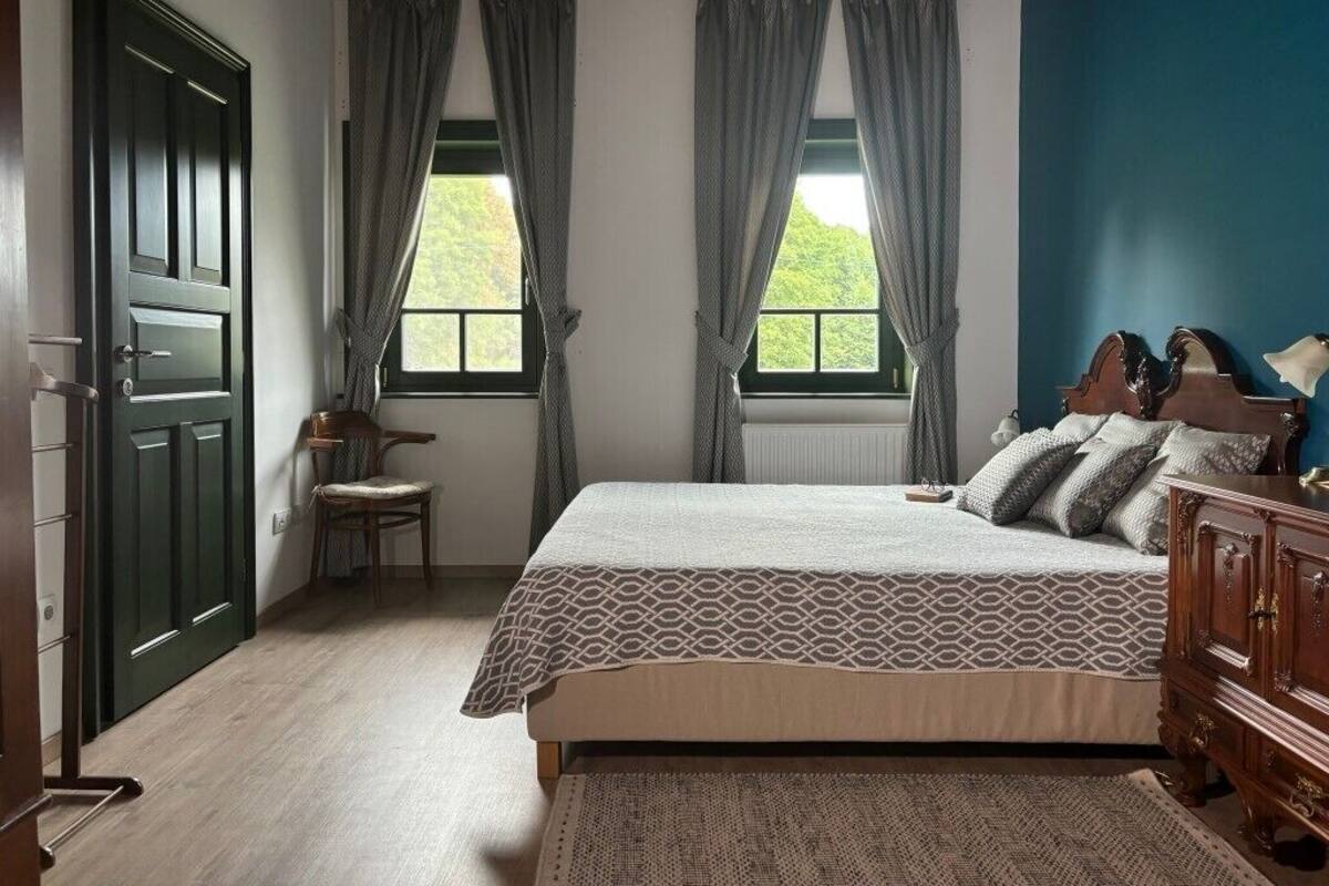 Quarto com cama, duas janelas com cortinas, uma cadeira, cômoda de madeira e uma porta.