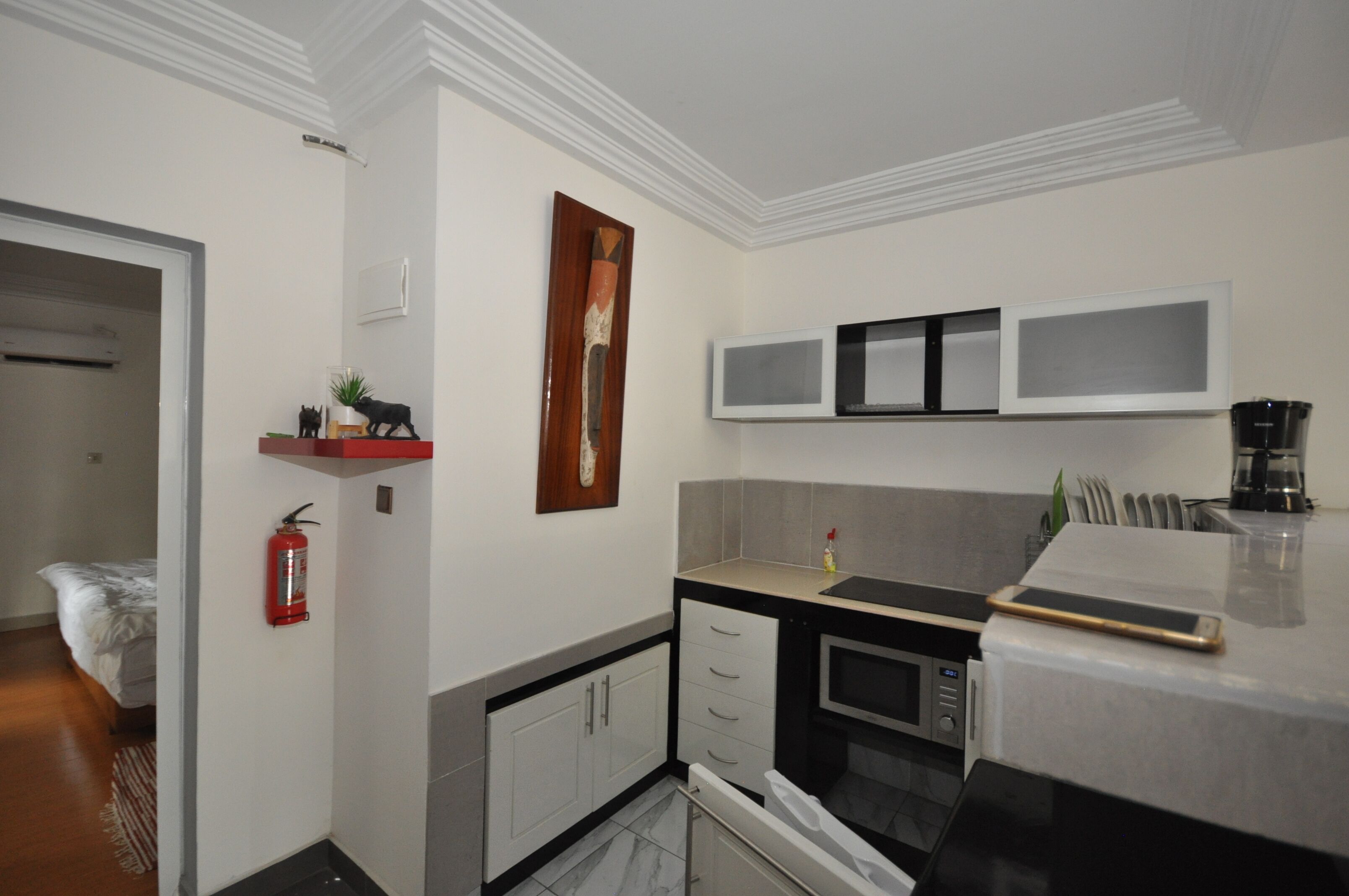 Uma cozinha compacta com micro-ondas, cafeteira e um balcão pequeno. Há um extintor de incêndio fixado na parede e uma prateleira com objetos decorativos.