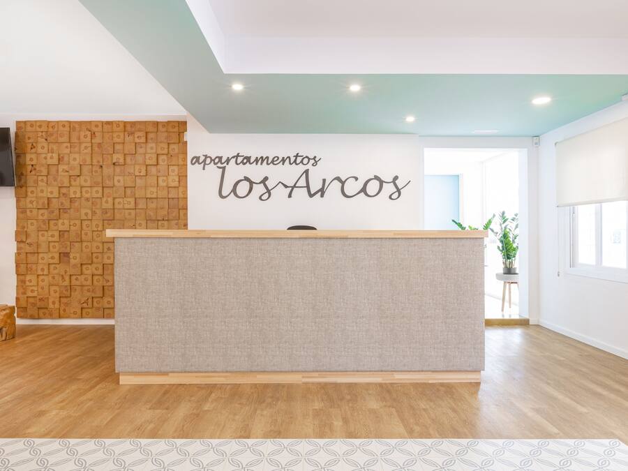 Uma área de recepção com parede de madeira, balcão de recepção cinza e uma placa que diz "apartamentos los Arcos".