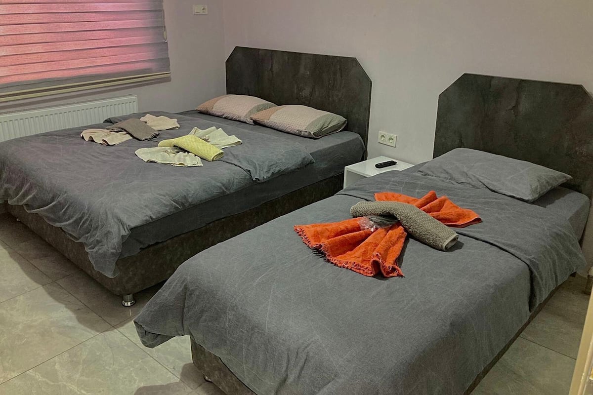 Quarto com duas camas, cada uma com roupa de cama e almofadas cinzas. Tem uma mesinha de cabeceira com um controle remoto.
