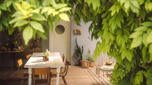 Uma área de refeições ao ar livre com uma mesa e cadeiras de madeira, cercada por plantas em vasos e uma planta pendente.