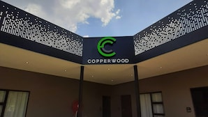 Um edifício moderno com a placa 'Copperwood', apresentando uma entrada central com galeria coberta e uma área gramada na frente.