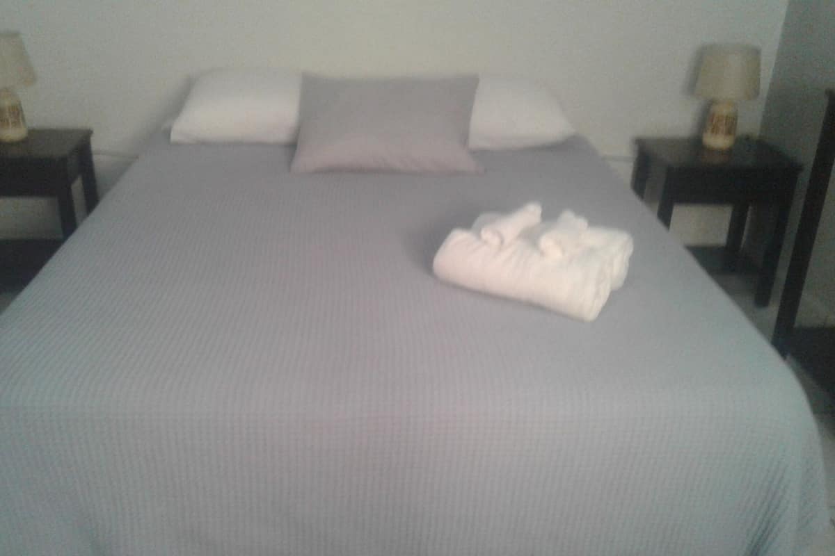 Uma cama com colcha cinza, toalha branca dobrada e duas mesas de cabeceira com abajures.