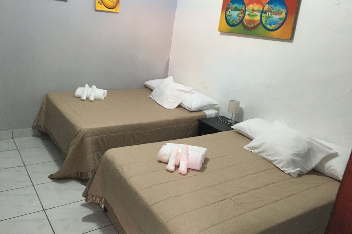 Quarto com duas camas, cada uma com colcha marrom e almofadas brancas. Há toalhas sobre as camas e um quadro com vasos na parede.