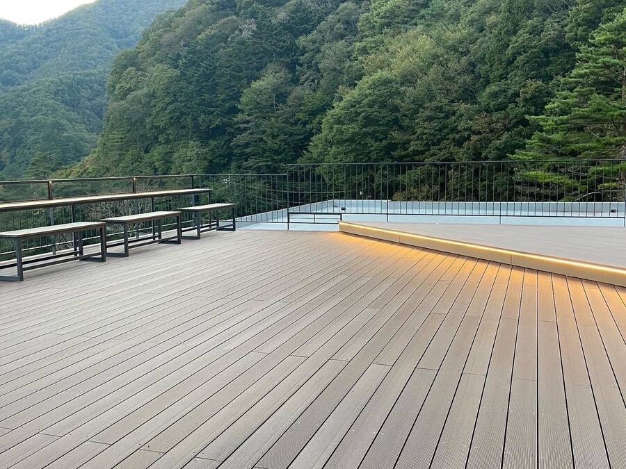 Um deck de madeira com bancos, cercado por guarda-corpo de metal e com vista para uma encosta arborizada.