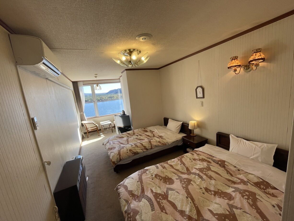 Quarto de hotel com duas camas, uma escrivaninha, uma cadeira, uma televisão e vista para o exterior.