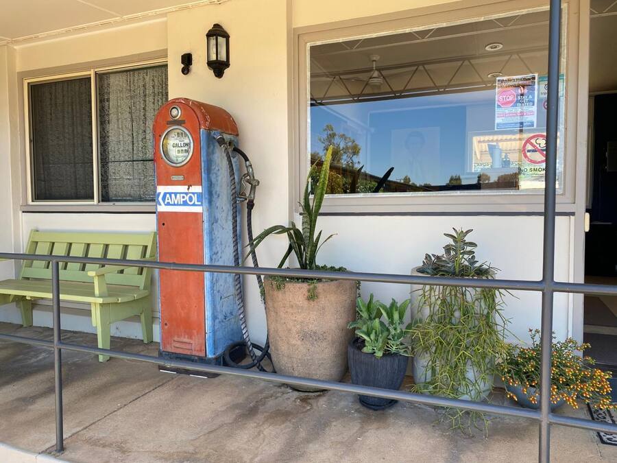 Uma bomba de gasolina AMPOL antiga em frente a um edifício com um banco verde e plantas em vasos.