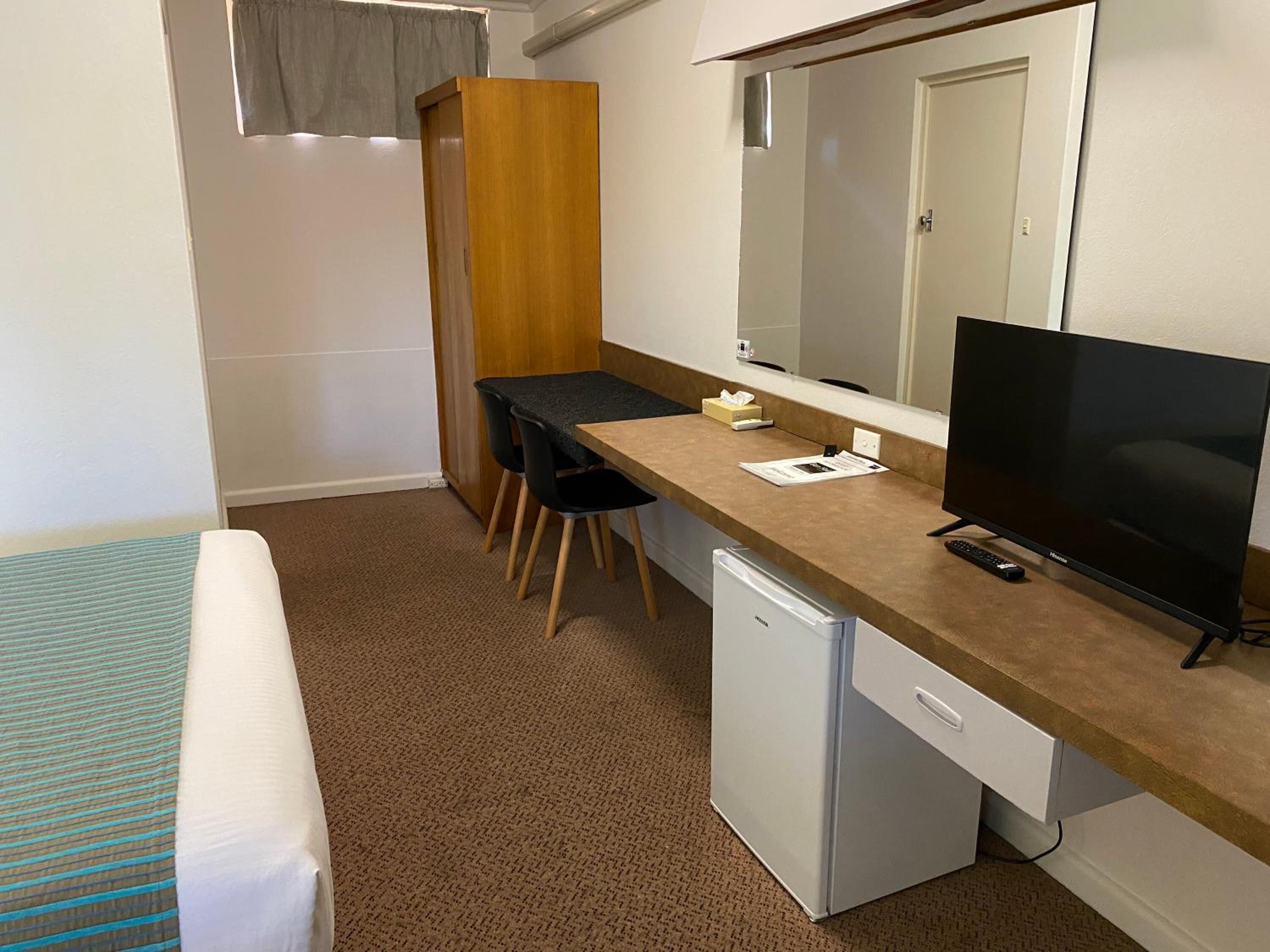 Quarto de hotel pequeno com uma cama de solteiro, uma escrivaninha com televisão, um frigorífico compacto e um guarda-roupa de madeira.