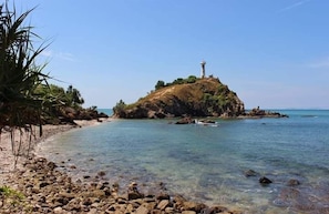 Uma praia rochosa com águas cristalinas, um farol numa pequena ilha e palmeiras.