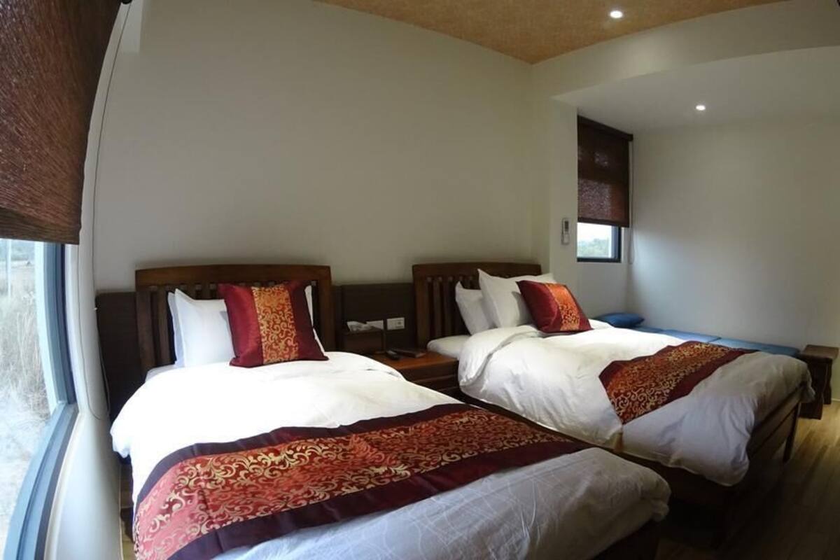 Quarto de hotel com duas camas, cada uma com cabeceira de madeira e roupa de cama branca. Há uma janela de um lado e uma parede de madeira do outro.