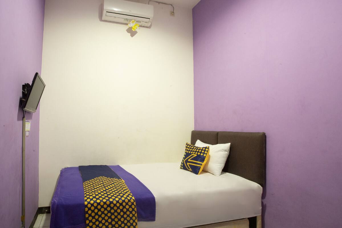 Uma cama de solteiro com cabeceira marrom, colchão branco e colcha estampada em roxo e amarelo.