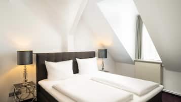 Quarto de hotel moderno com uma cama grande, duas mesas de cabeceira com abajures, uma janela com cortinas e teto inclinado.