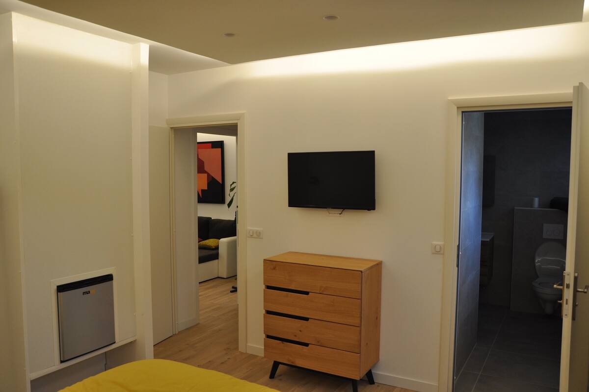 Quarto com cama, cómoda de madeira, televisão na parede e banheiro visível por uma porta aberta.