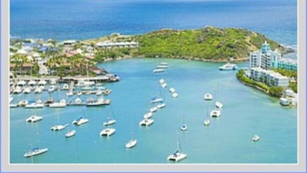 Uma marina costeira com vários barcos atracados, uma pequena ilha com edifícios e vista clara do oceano.
