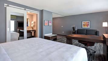 Quarto de hotel com cama grande, escrivaninha, cadeira, televisão e banheiro com espelho, além de obras de arte nas paredes.
