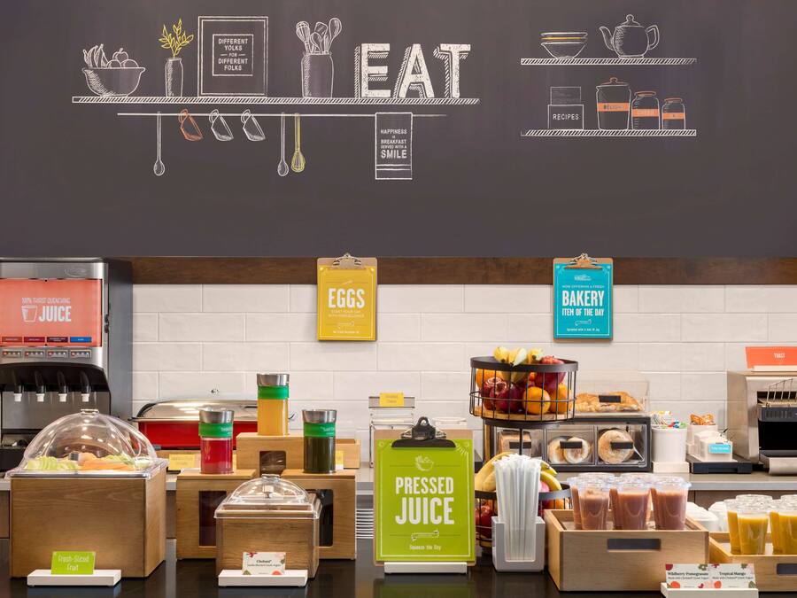 Uma mesa de pequeno-almoço com um menu num quadro negro, um dispensador de sumo e diversas opções de comida.