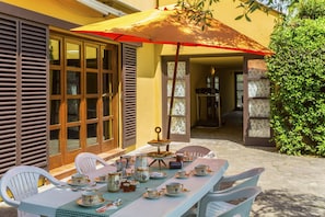Uma mesa posta para o chá, com cadeiras brancas e um guarda-sol vermelho, posicionada em frente a um edifício com persianas de madeira e um pátio com azulejos.