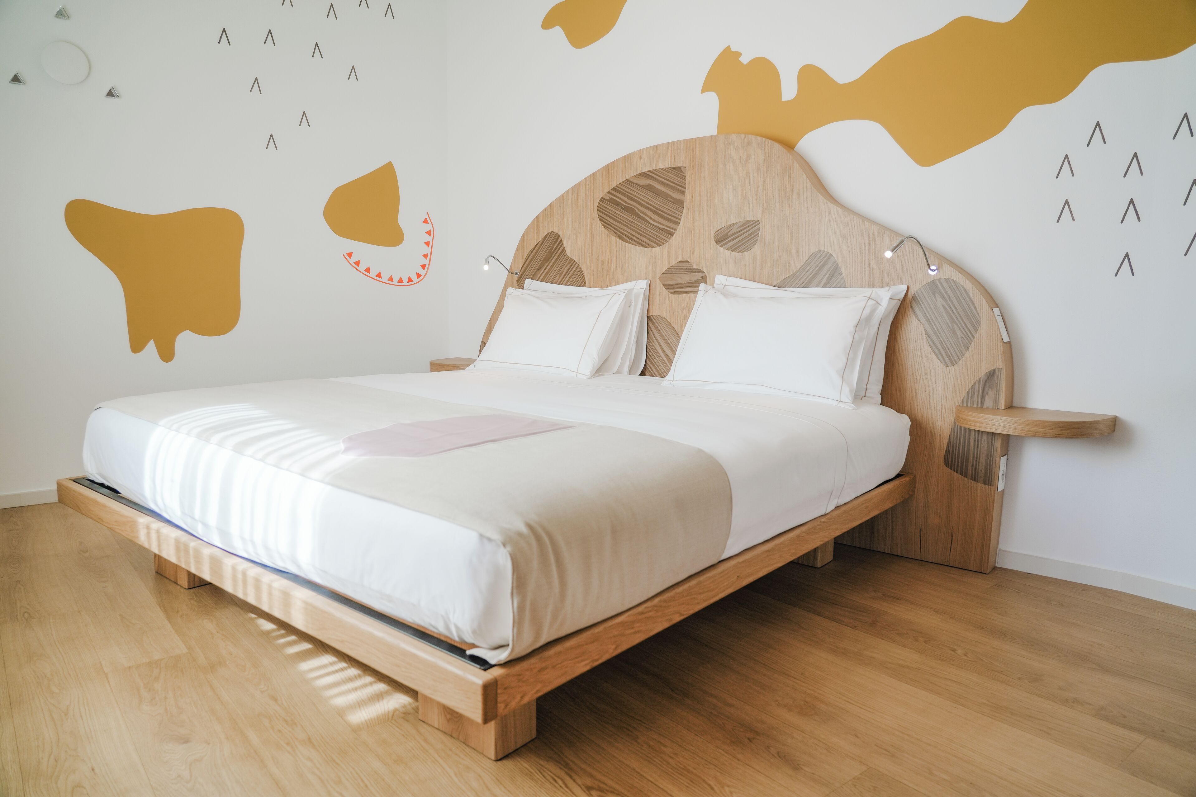 Quarto moderno com estrutura de cama de madeira, roupa de cama branca e parede decorada com arte abstrata de animais e formas geométricas.