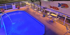 Uma piscina ao ar livre com uma fonte, cercada por cadeiras de metal e plantas em vasos.