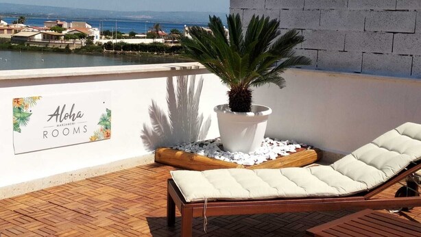 Uma varanda com uma cadeira de lounge, um vaso com planta e uma placa com a inscrição 'Aloha Marzamemi Rooms'.