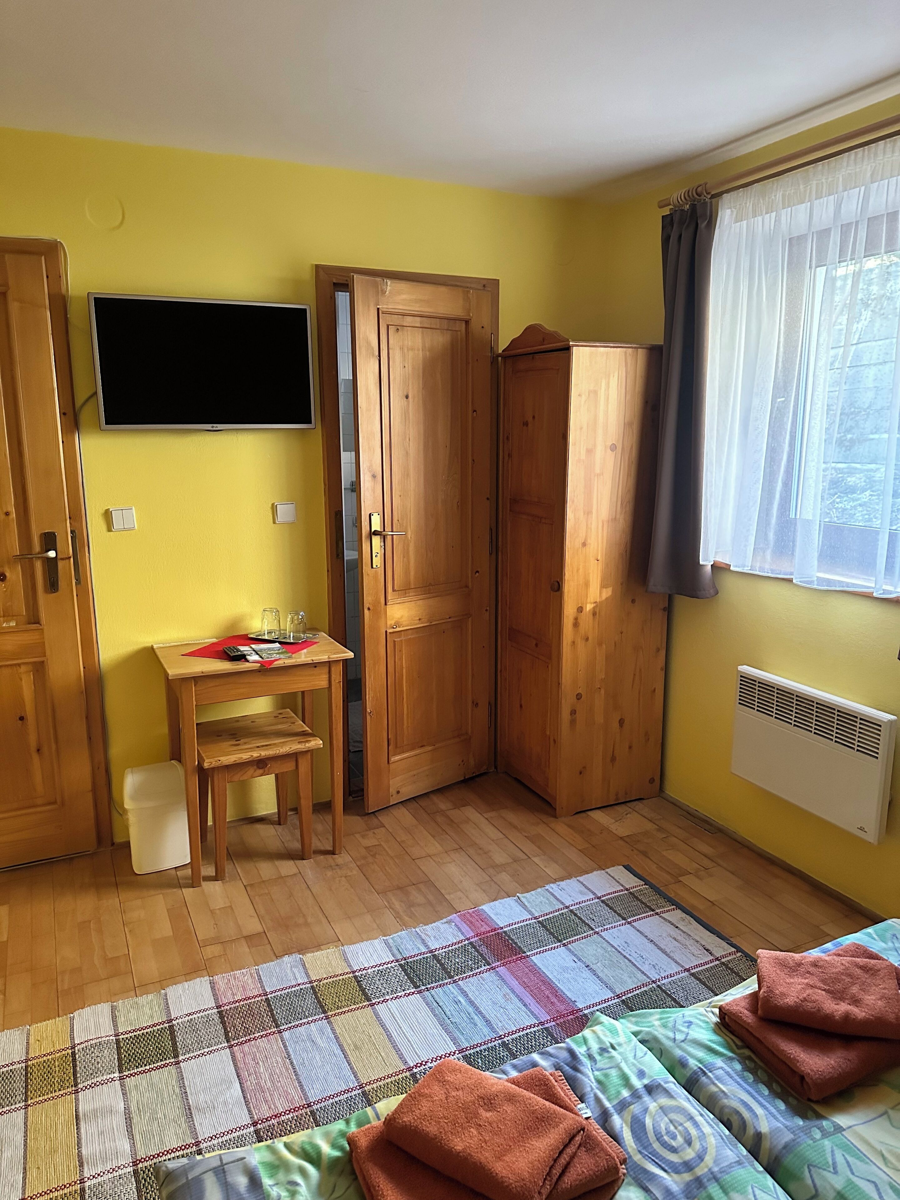 Quarto com cama, guarda-roupa de madeira, uma pequena mesa com copos e televisão fixada na parede.