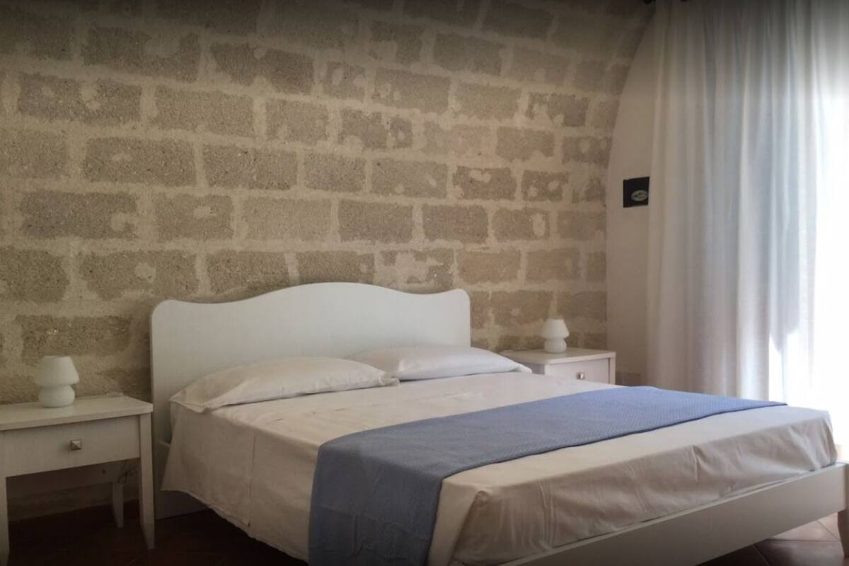 Quarto com parede de tijolos, cama branca com manta azul, duas mesas de cabeceira com luminárias e uma janela com cortinas.