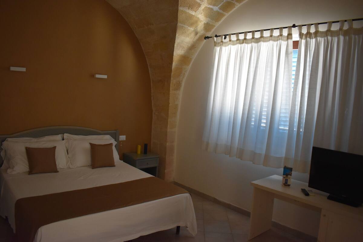 Quarto com cama, mesa de cabeceira, televisão e janela com cortinas.