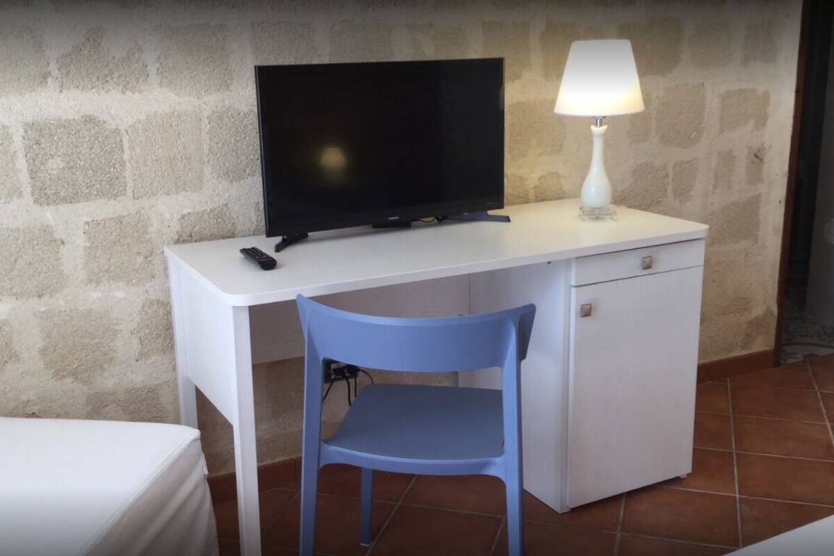 Um quarto pequeno com uma escrivaninha branca, uma televisão preta, uma cadeira azul e um abajur branco.