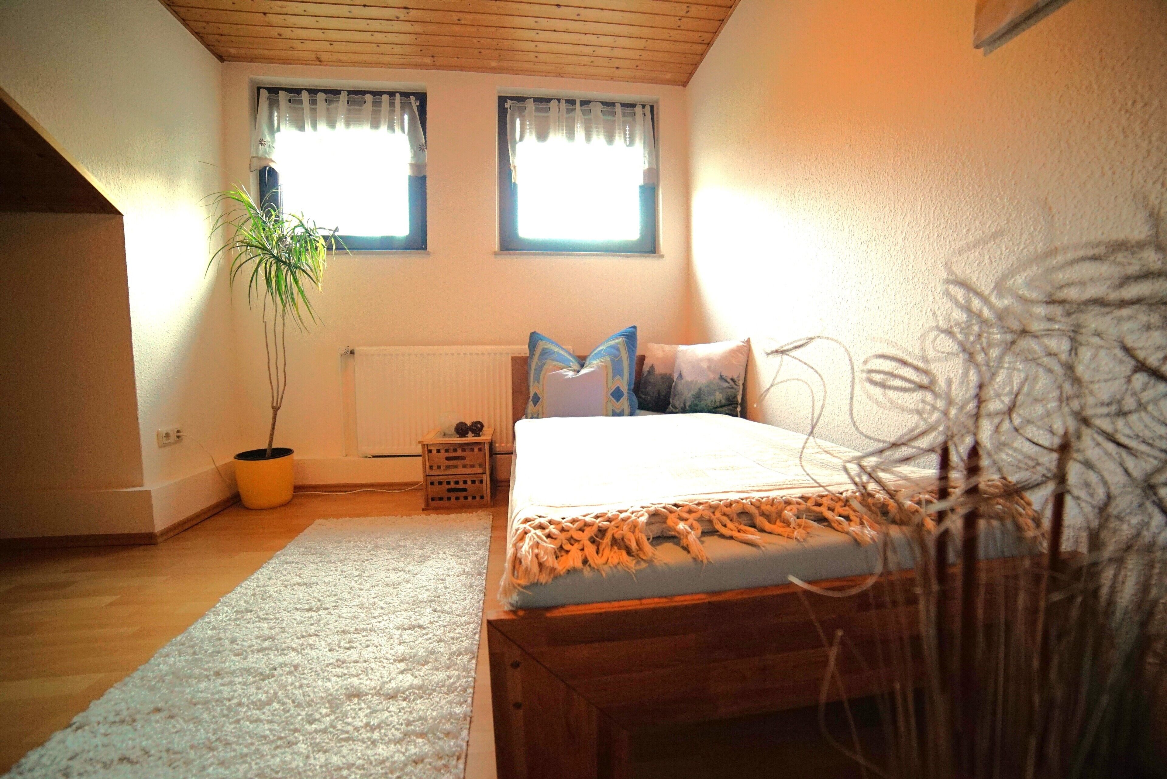 Quarto com teto de madeira, cama com colchão branco e almofadas azuis, radiador branco, planta em vaso e um pequeno baú de madeira.