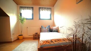 Quarto com teto de madeira, cama com colchão branco e almofadas azuis, radiador branco, planta em vaso e um pequeno baú de madeira.