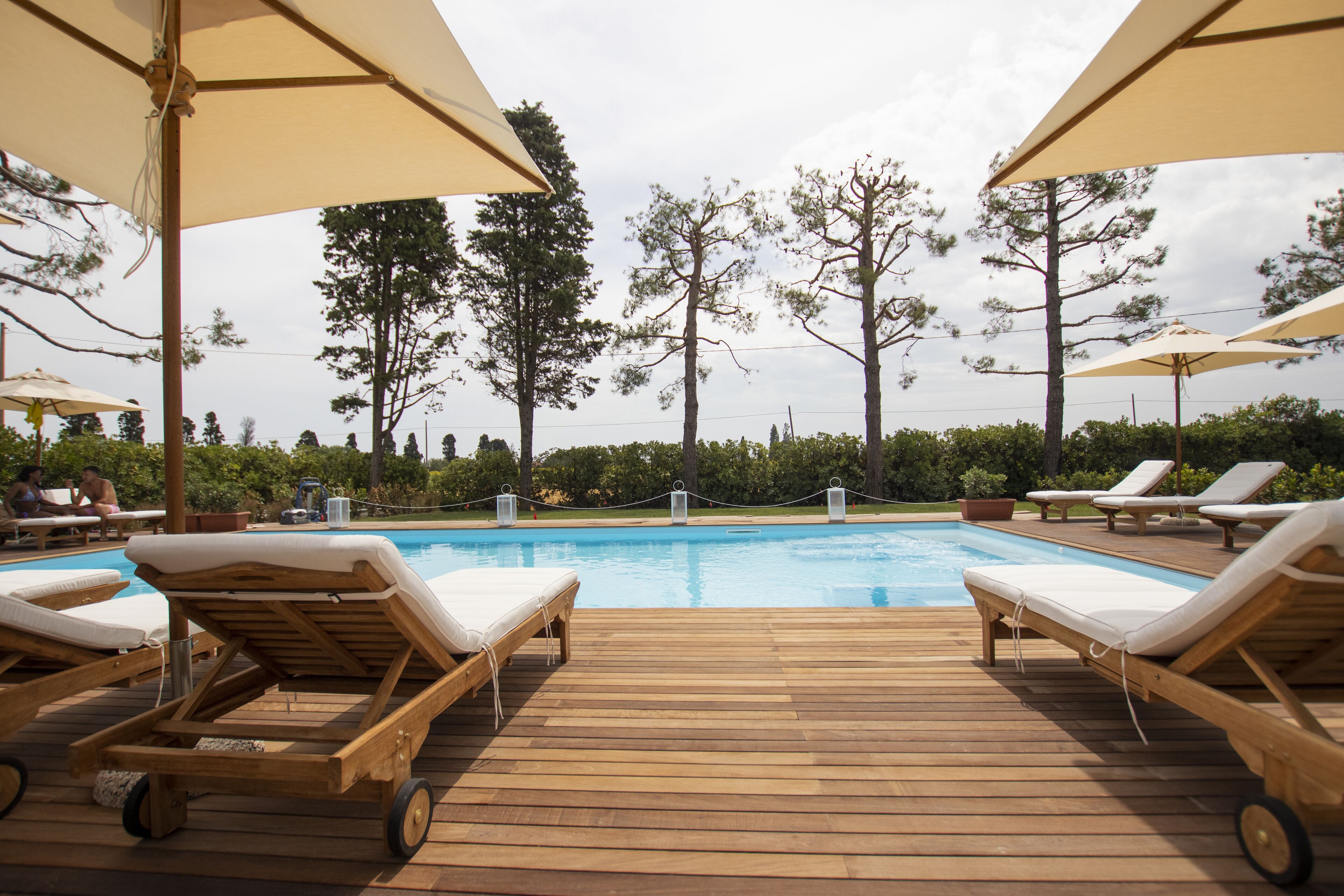 Um deck de madeira com cadeiras de lounge e guarda-sóis com vista para uma piscina, cercado por árvores.