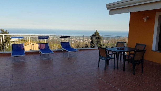 Terraço com espreguiçadeiras azuis e uma mesa com cadeiras, com vista para a paisagem urbana e o mar.