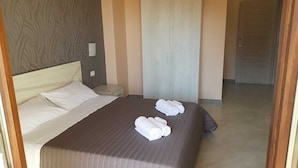 Cama bem arrumada com lençóis brancos e toalhas dobradas, cabeceira de madeira, mesa de cabeceira e parede lisa com interruptor de luz.