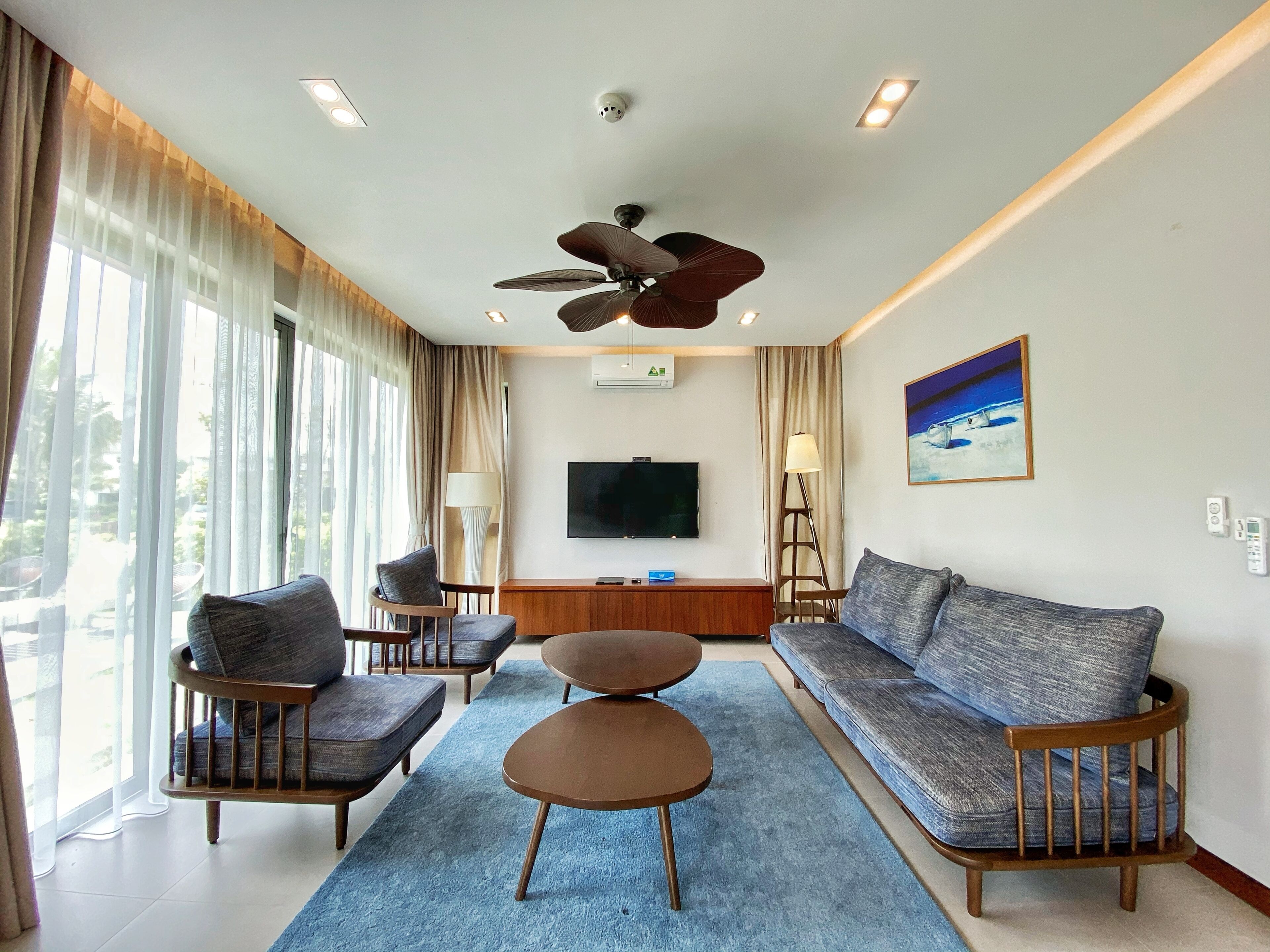 Quarto de hotel moderno com sofá, duas poltronas, mesa de centro, ventoinha no teto, televisão de tela plana e um quadro na parede.