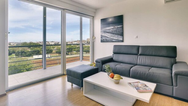 Uma sala de estar moderna com um sofá cinzento, uma mesa de centro branca com frutas, e uma grande janela com vista para a cidade.