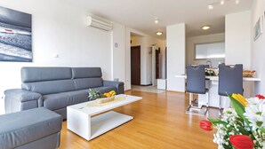 Uma sala de estar moderna com um sofá cinzento, uma mesa de centro branca com frutas, e uma área de cozinha com duas cadeiras e uma pia.