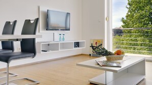 Uma sala de estar moderna com televisão de tela plana na parede, mesa de centro branca com uma cesta de frutas, e uma área de estar com sofá e poltronas.