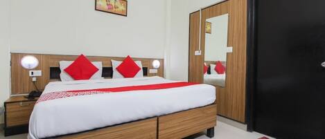Quarto de hotel com cama de madeira, almofadas vermelhas, mesa de cabeceira com luminária, espelho e guarda-roupa.