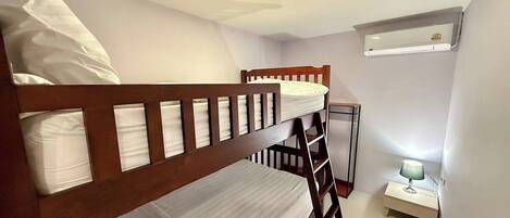 Quarto com beliche, mesa de cabeceira com abajur e aparelho de ar condicionado na parede.