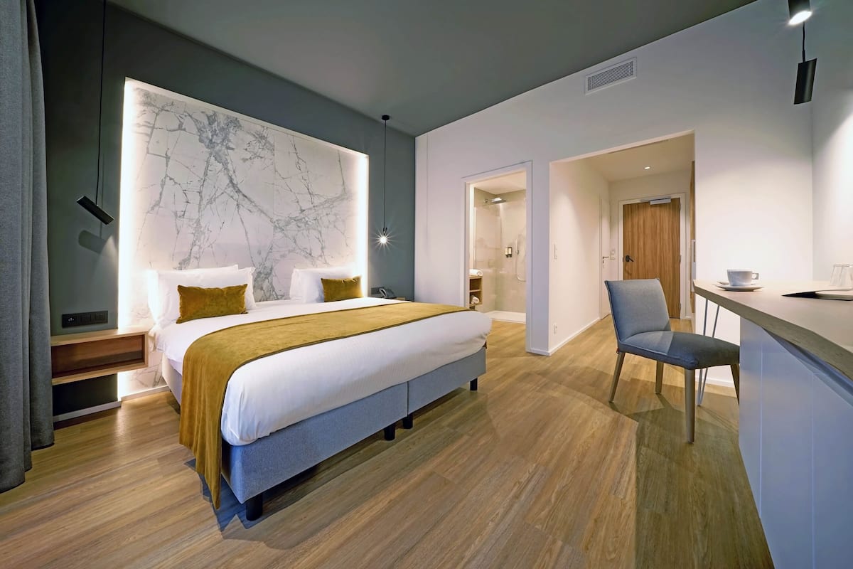 Quarto moderno com uma cama grande, uma mesa de trabalho e uma cadeira. Um banheiro é visível através de uma porta aberta.