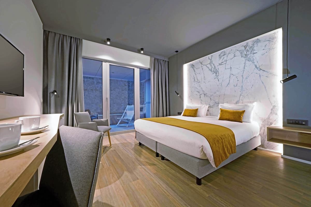 Quarto de hotel moderno com uma cama grande, uma escrivaninha com cadeira, uma televisão e vista para o exterior.