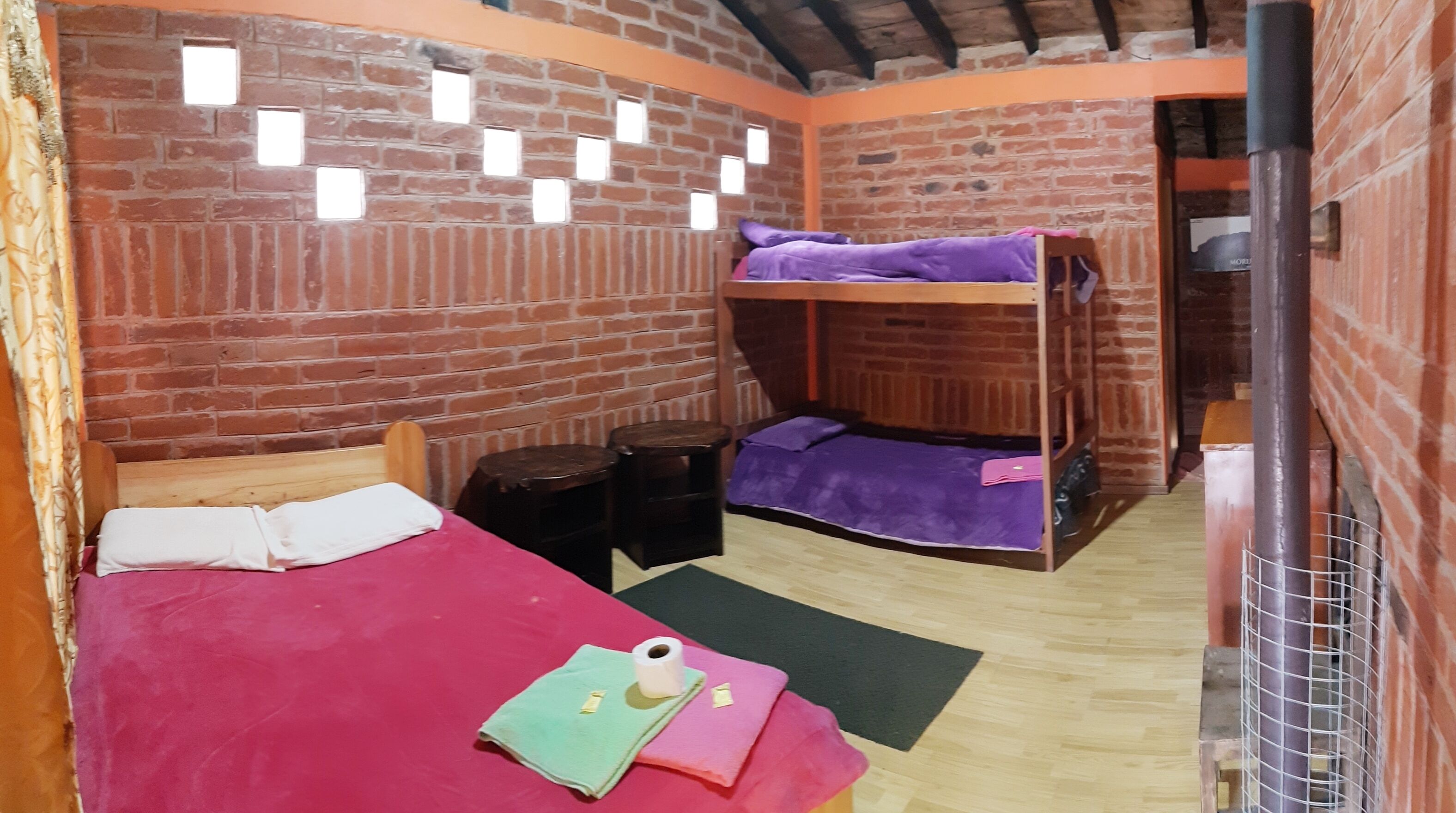 Quarto com parede de tijolos, uma cama de solteiro, uma cama beliche, uma mesinha e uma cadeira.